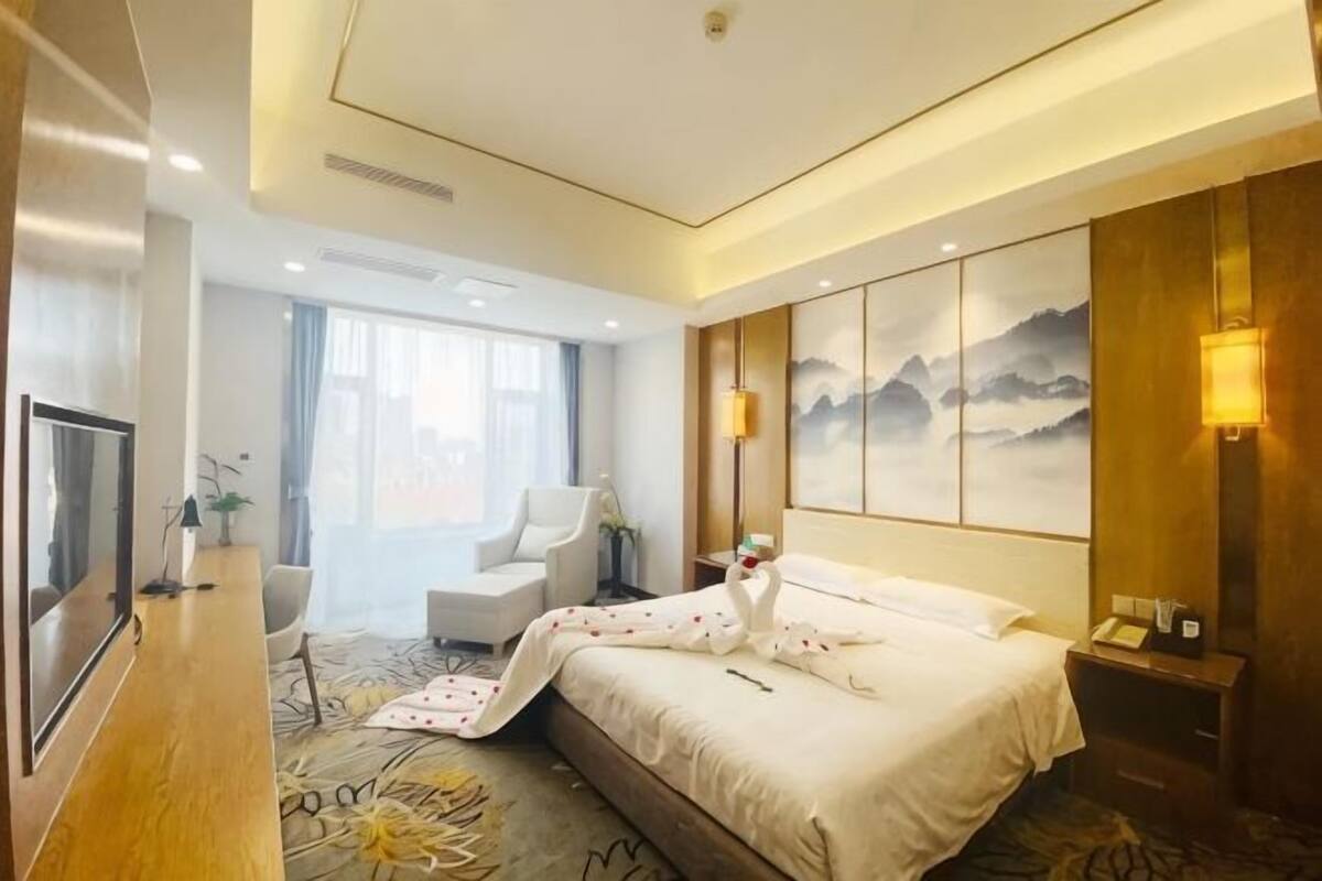 Quarto de hotel com cama, escrivaninha, cadeira, televisão e um quadro na parede.