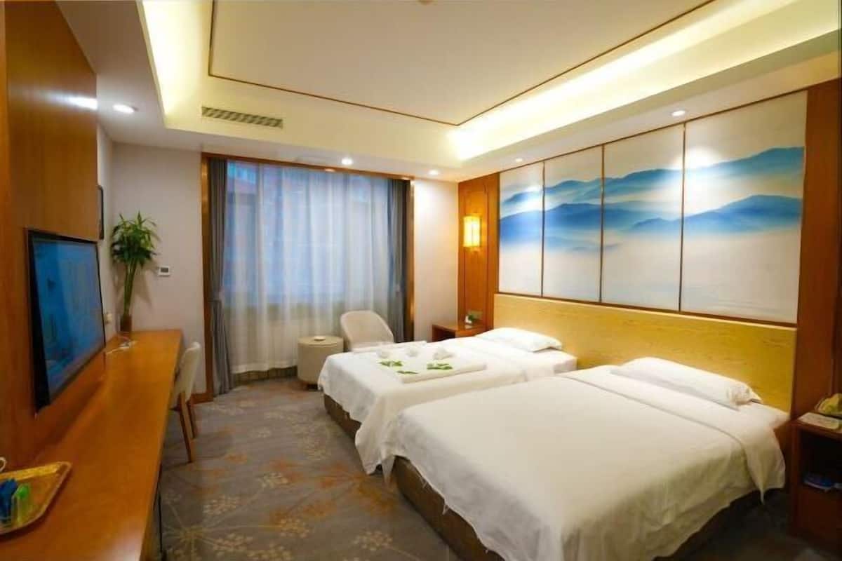 Quarto de hotel com duas camas, uma escrivaninha, uma cadeira, uma televisão e um quadro na parede.