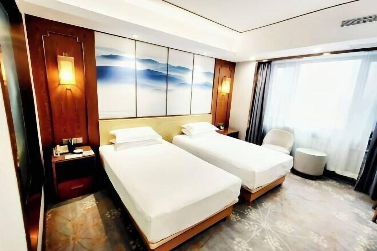 Quarto de hotel com duas camas de solteiro, uma pequena mesa de cabeceira, uma cadeira e uma janela grande com cortinas.
