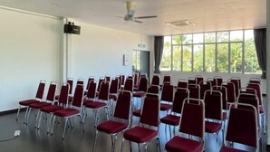 Uma sala de conferências com fileiras de cadeiras vermelhas, um ventilador de teto e amplas janelas com vista para o verde.