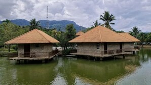 Duas casas flutuantes de madeira com telhados de palha em um lago calmo, rodeadas por palmeiras e montanhas.