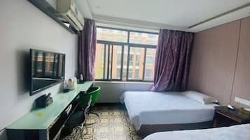 Quarto de hotel com duas camas, uma escrivaninha com televisão de tela plana, uma cadeira verde e uma janela com cortinas.