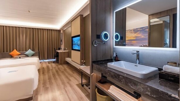 Quarto de hotel moderno com uma cama grande, televisão, banheiro com espelho amplo e piso de madeira.