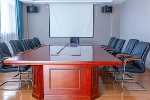 Uma sala de conferências com uma mesa comprida, cadeiras pretas, tela de projetor e quadro branco.