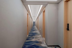 Corredor de hotel com carpete estampado, portas de madeira e luminária no teto.