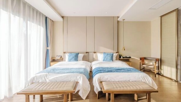 Quarto de hotel com duas camas, uma escrivaninha, uma cadeira e uma janela grande com cortinas.