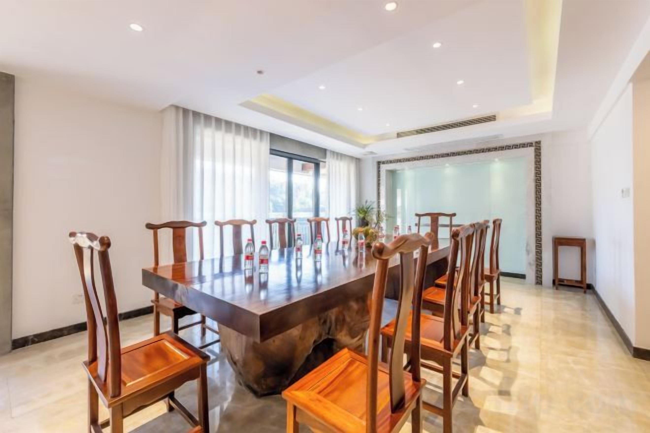 Sala de conferência com uma mesa retangular grande rodeada por cadeiras de madeira, uma divisória de vidro e uma janela com cortinas.