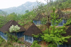 Edifícios em estilo tradicional com telhados de palha, rodeados por vegetação exuberante e colinas ao fundo.