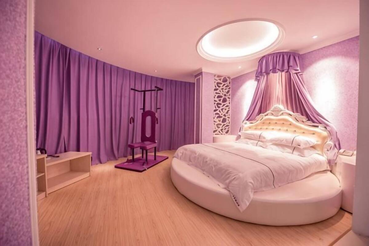 Uma cama redonda com dossel, um equipamento de exercício vertical e um pequeno armário em um quarto com cortinas roxas e parede estampada.