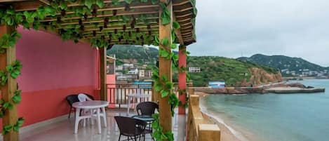 Uma área de estar ao ar livre com pérgola, cadeiras e vista para o mar e colinas.