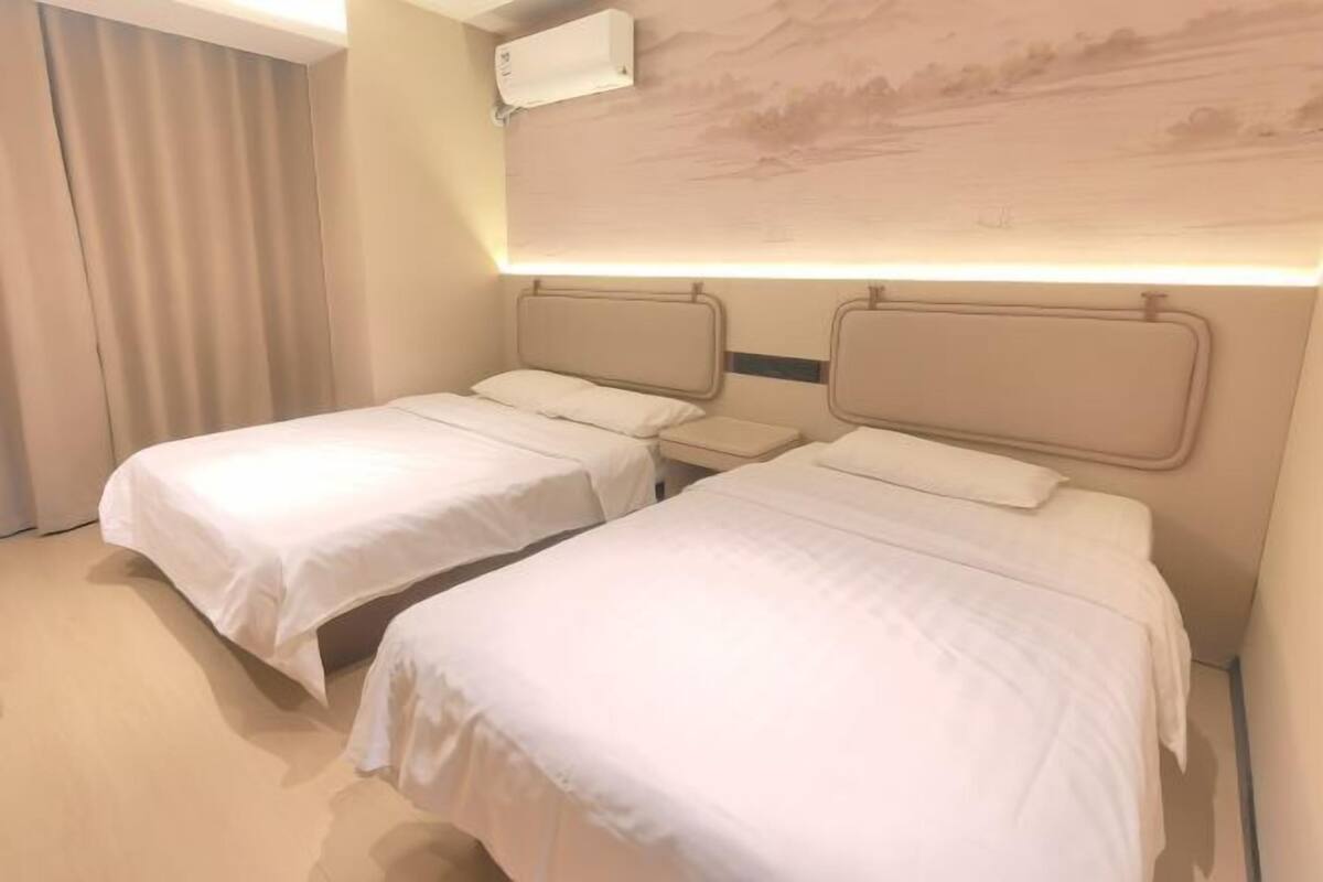 Quarto de hotel com duas camas, cabeceira com luminária e ar condicionado na parede, além de cortina.