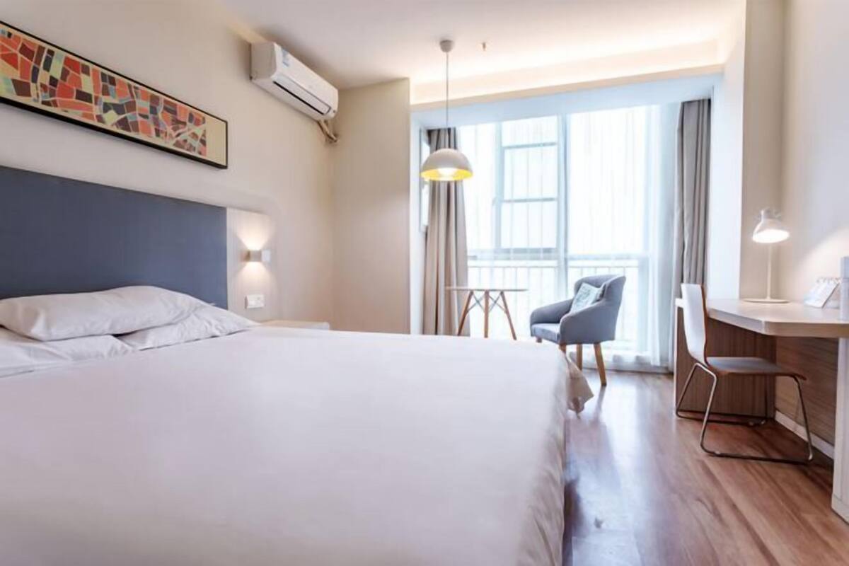 Quarto de hotel com cama, escrivaninha, cadeira e janelas amplas.
