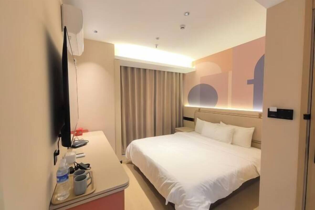 Quarto de hotel com cama, mesa de cabeceira, televisão e cortinas.
