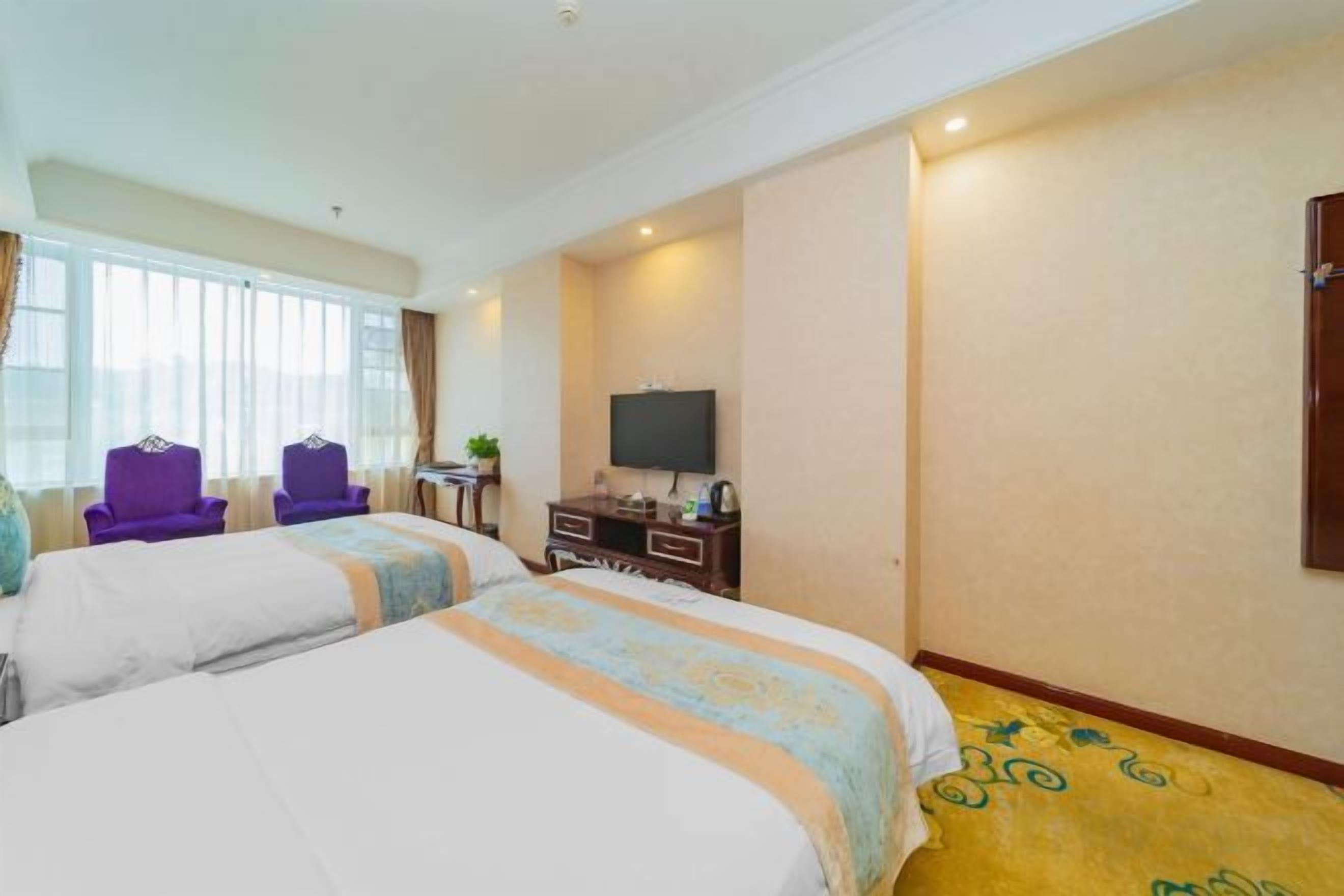 Quarto de hotel com duas camas, televisão, escrivaninha e janela com cortinas.