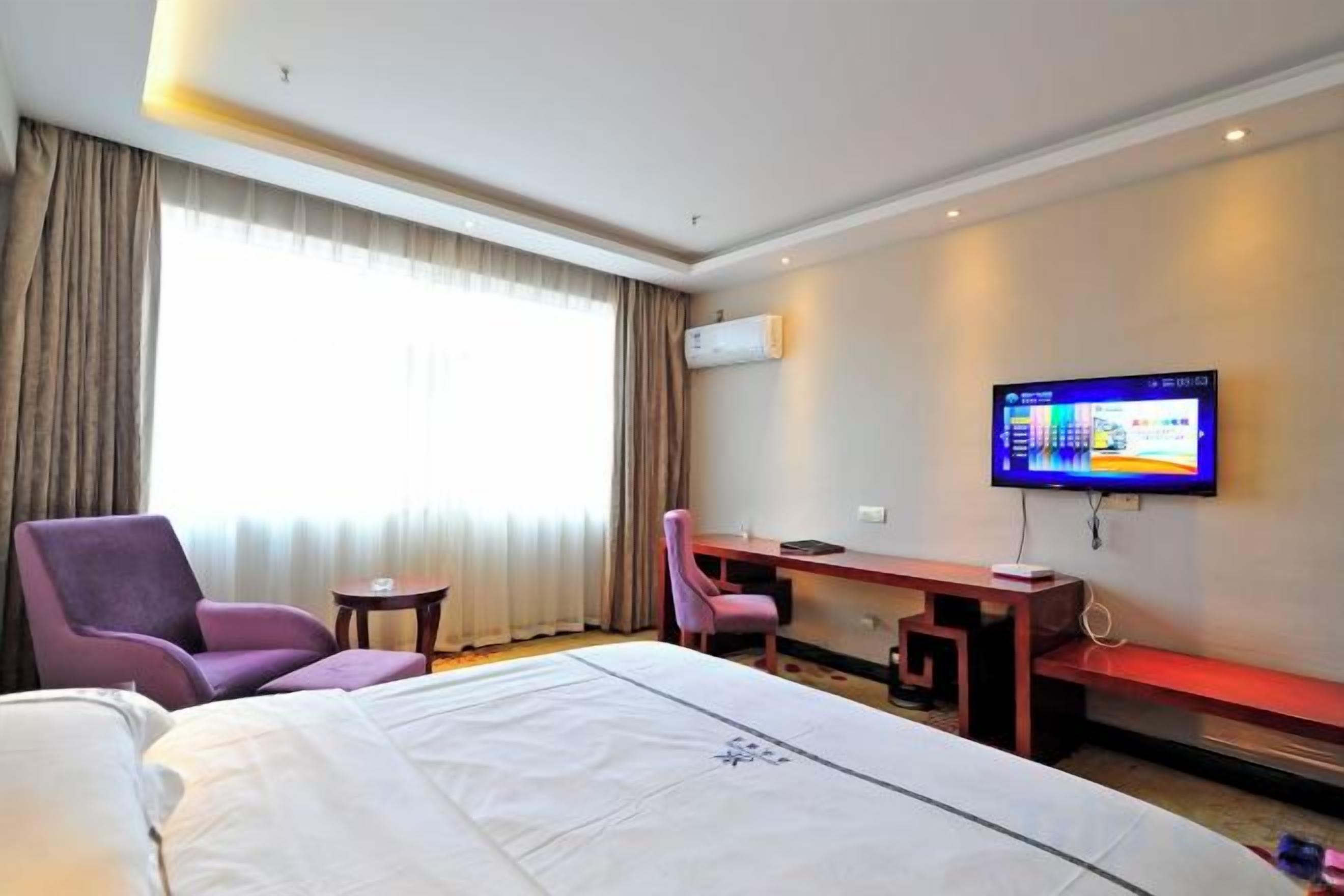 Quarto de hotel com cama, escrivaninha, cadeira, televisão e janela com cortinas.