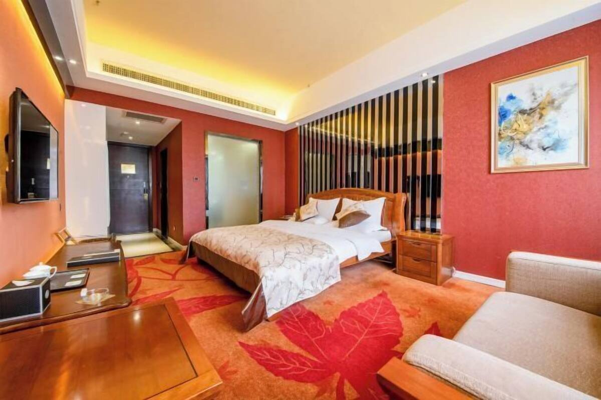 Quarto de hotel com cama, escrivaninha, cadeira, televisão e um quadro na parede.