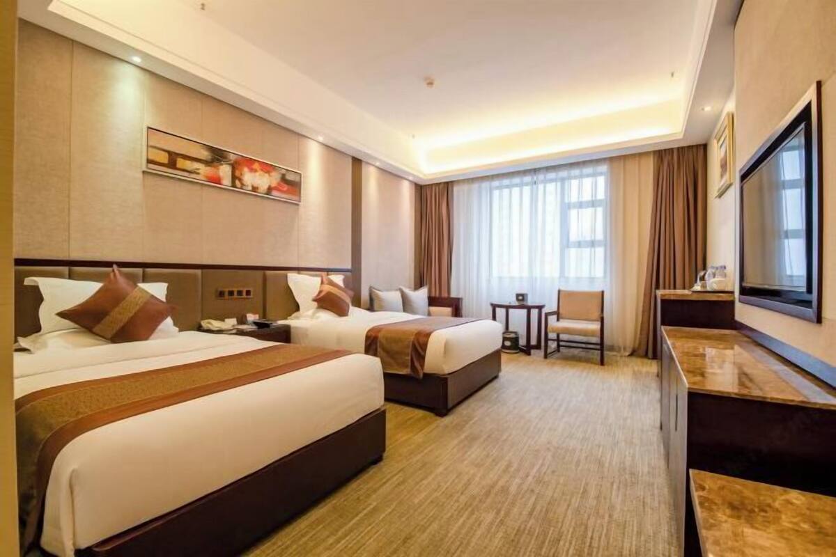 Quarto de hotel com duas camas, uma mesinha, uma cadeira, uma televisão e um quadro na parede.