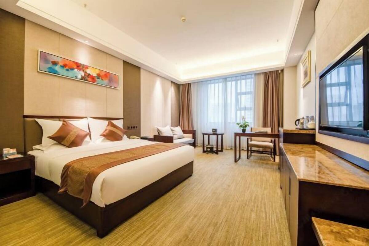 Quarto de hotel com uma cama grande, uma escrivaninha, uma cadeira, uma mesinha e uma televisão.