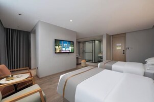 Quarto de hotel com duas camas, uma televisão fixada na parede, uma mesinha com uma cadeira e uma porta de vidro para o banheiro.
