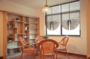 Quarto com uma mesa de jantar, quatro cadeiras de vime, uma estante de madeira e uma janela grande com ventilador.