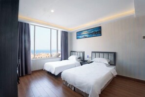Quarto de hotel com duas camas, um quadro na parede e vista da cidade através de grandes janelas.