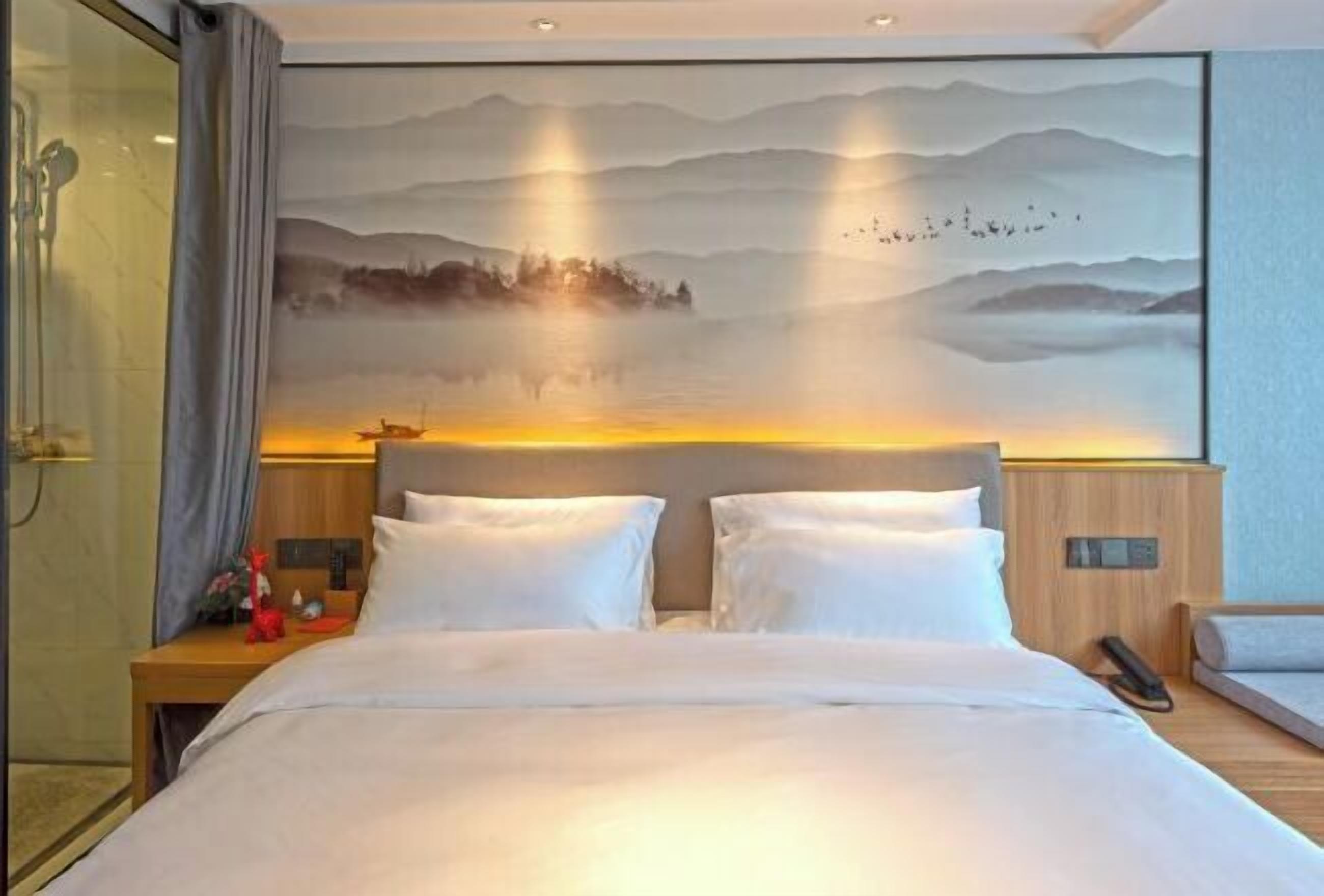 Quarto de hotel com uma cama grande, mesa de cabeceira com abajur, telefone fixado na parede e uma pintura grande de uma paisagem.
