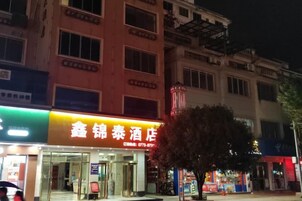 Um edifício com uma placa bem iluminada, uma fachada com entrada visível e uma árvore na frente.