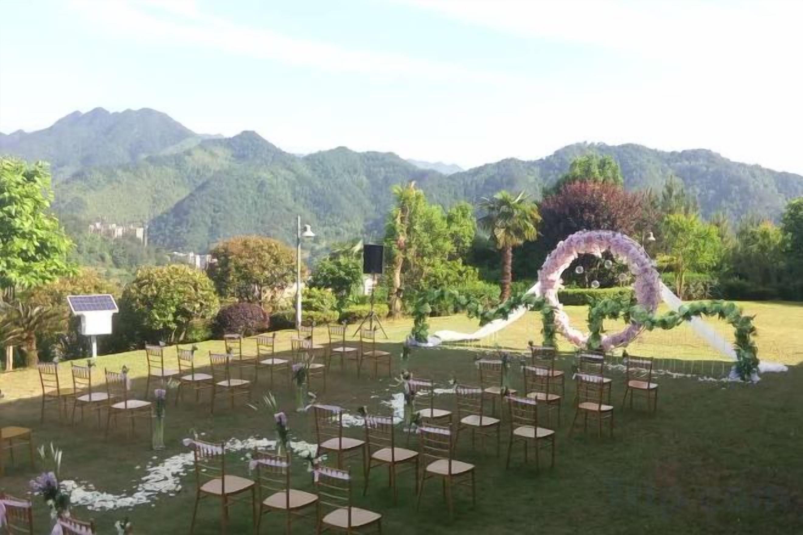 Espaço para casamento ao ar livre com disposição circular de assentos, um arco decorativo e montanhas ao fundo.
