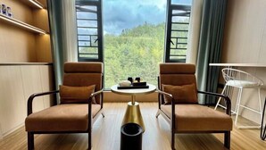 Quarto moderno com duas poltronas castanhas, uma mesa redonda pequena e vista para uma floresta através de grandes janelas.