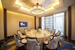 Sala de conferência com mesa redonda, cadeiras, lustre, televisão e uma janela ampla.