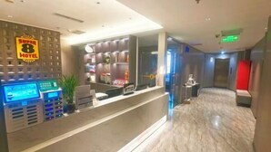 Uma área de recepção de hotel com design moderno, incluindo um balcão de atendimento, um totem de check-in digital e uma placa fixada na parede.