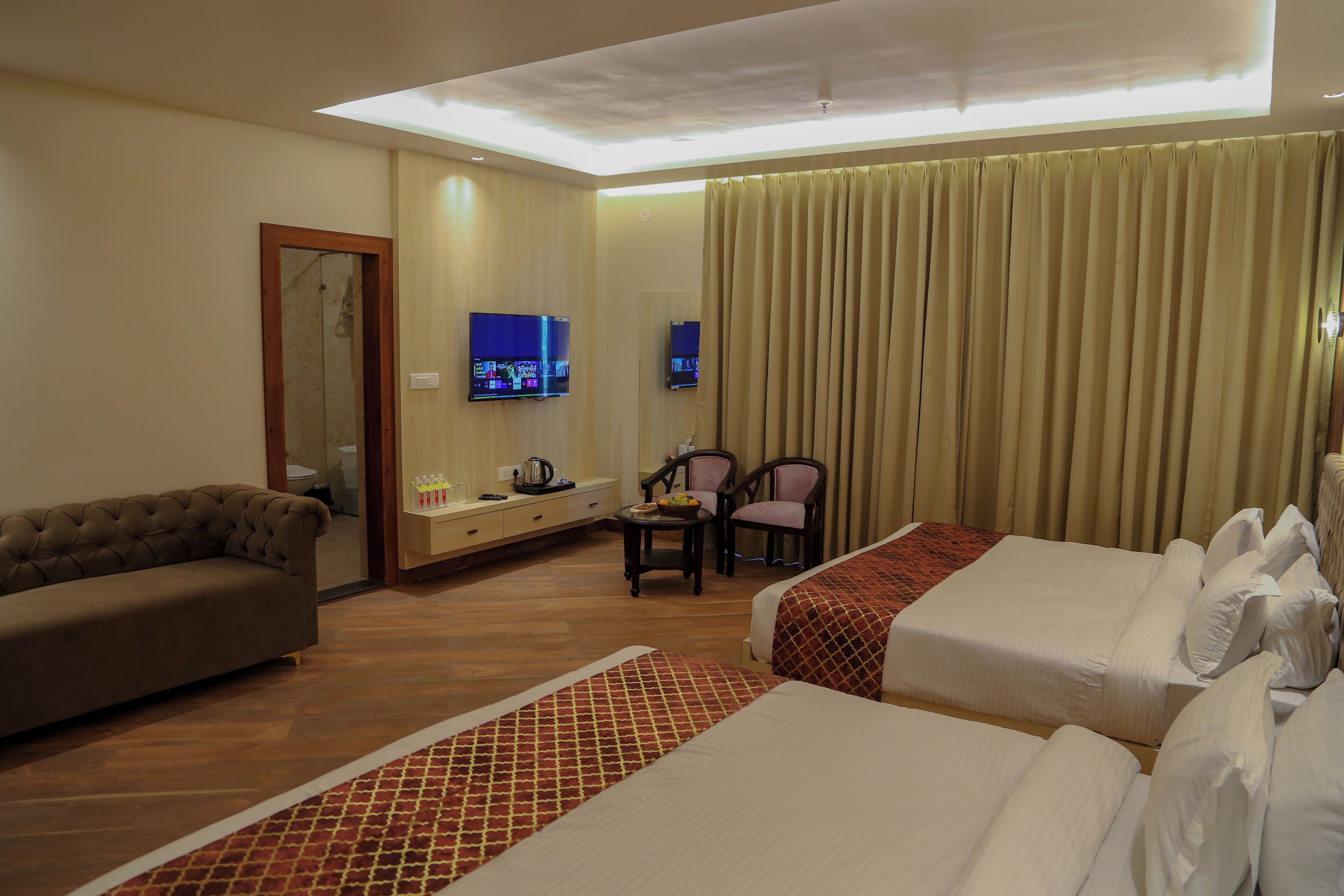 Quarto de hotel com duas camas, um sofá, uma TV e uma escrivaninha com cadeiras.