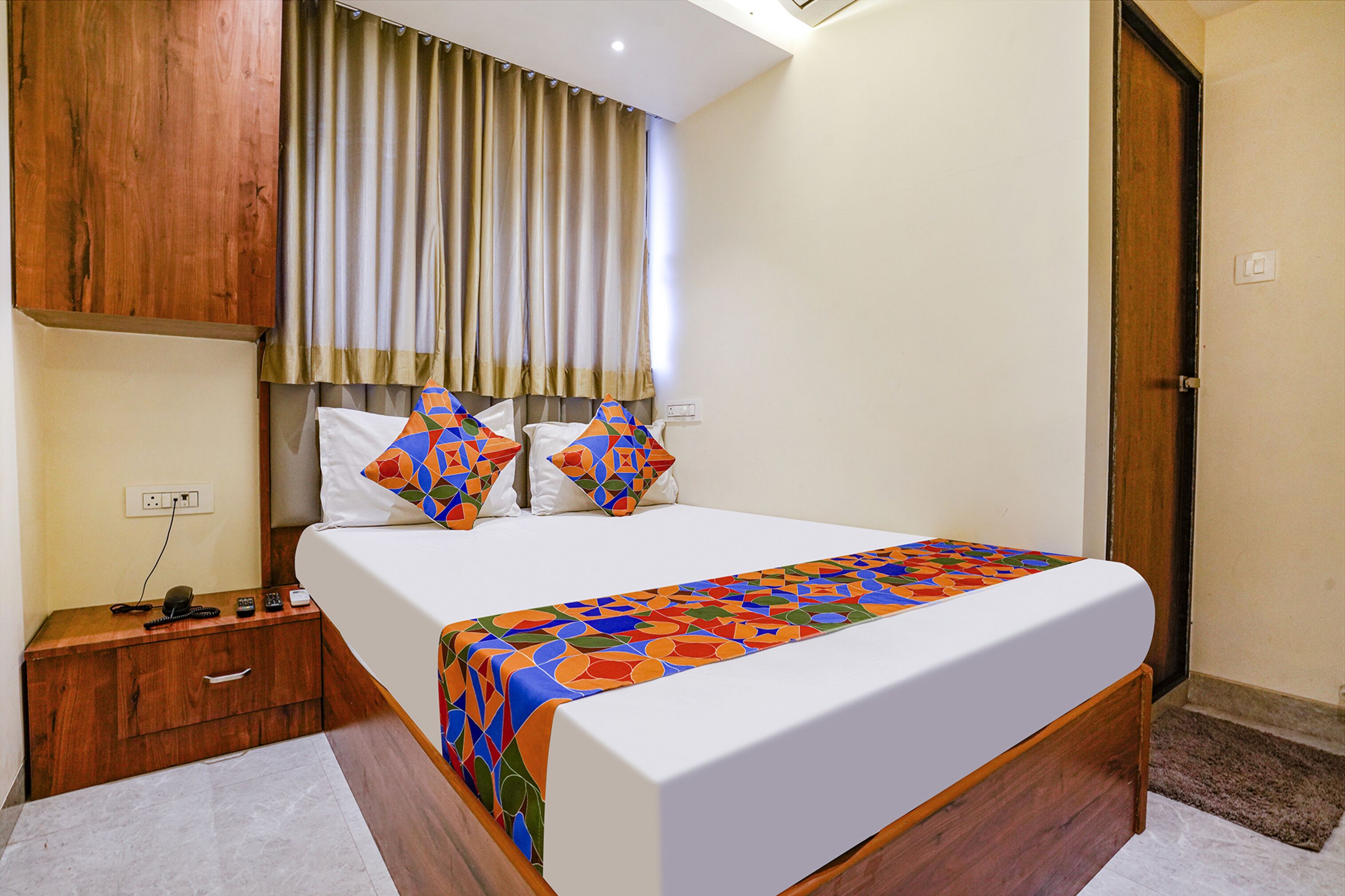 Cama bem arrumada com colcha colorida, mesa de cabeceira de madeira com telefone e controle remoto, e janela com cortinas.