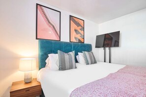 Uma cama bem arrumada com cabeceira verde-água, uma luminária de mesa e uma TV fixada na parede.