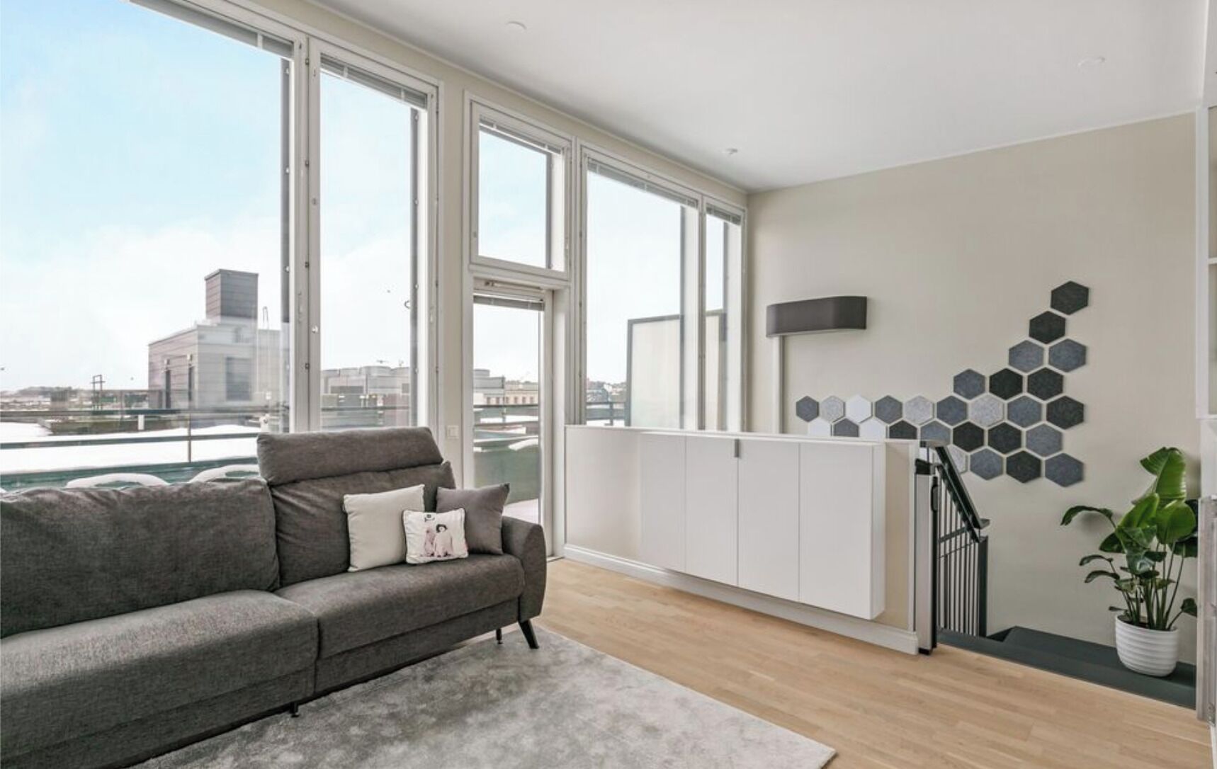Uma sala de estar moderna com um sofá cinzento, janelas amplas, um radiador branco e uma decoração de parede hexagonal.