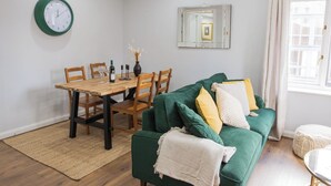Sala de estar com sofá verde, mesa de jantar de madeira e cadeiras. Há um relógio na parede e um espelho acima do sofá.