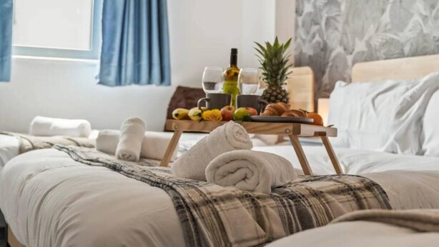 Uma cama bem arrumada com um cobertor xadrez, uma bandeja com frutas, uma garrafa e uma abacaxi em uma mesa de cabeceira.