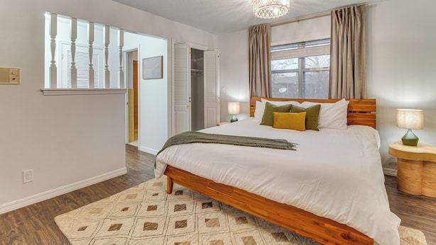 Quarto com cama de madeira, mesa de cabeceira, luminária, janela com cortinas e porta que dá para outro cômodo.