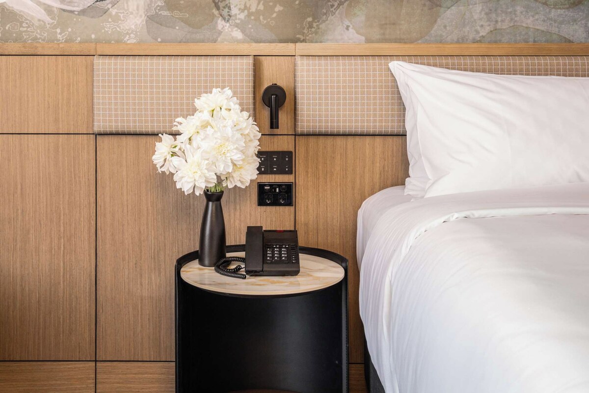 Quarto de hotel com cabeceira de madeira, mesa de cabeceira com telefone e um vaso de flores, e cama com roupa de cama branca.