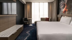 Quarto de hotel com cama, televisão, escrivaninha, cadeira, mesinha e um quadro na parede.