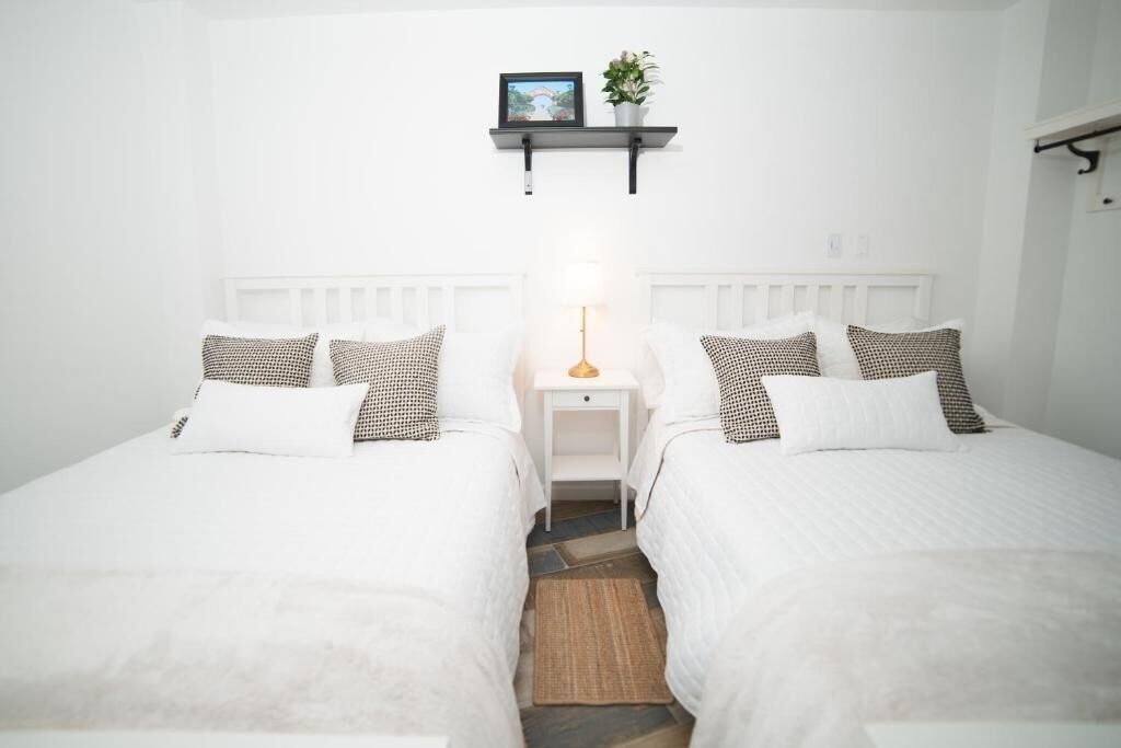 Quarto duplo com duas camas, roupa de cama branca, mesa de cabeceira com abajur, pequena estante com um quadro e uma planta, e cabeceira branca.