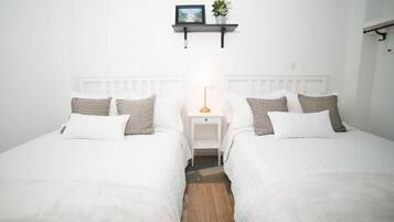 Quarto duplo com duas camas, roupa de cama branca, mesa de cabeceira com abajur, pequena estante com um quadro e uma planta, e cabeceira branca.