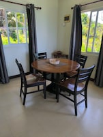 Uma mesa de jantar redonda de madeira com seis cadeiras, uma mesinha lateral e uma janela com cortinas.