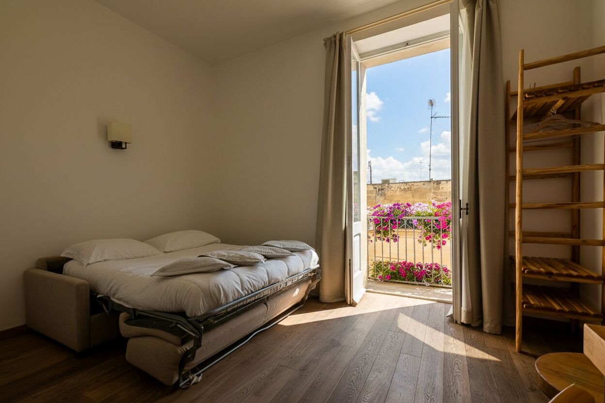 Quarto com cama, uma mesinha, estante e vista para uma varanda florida.