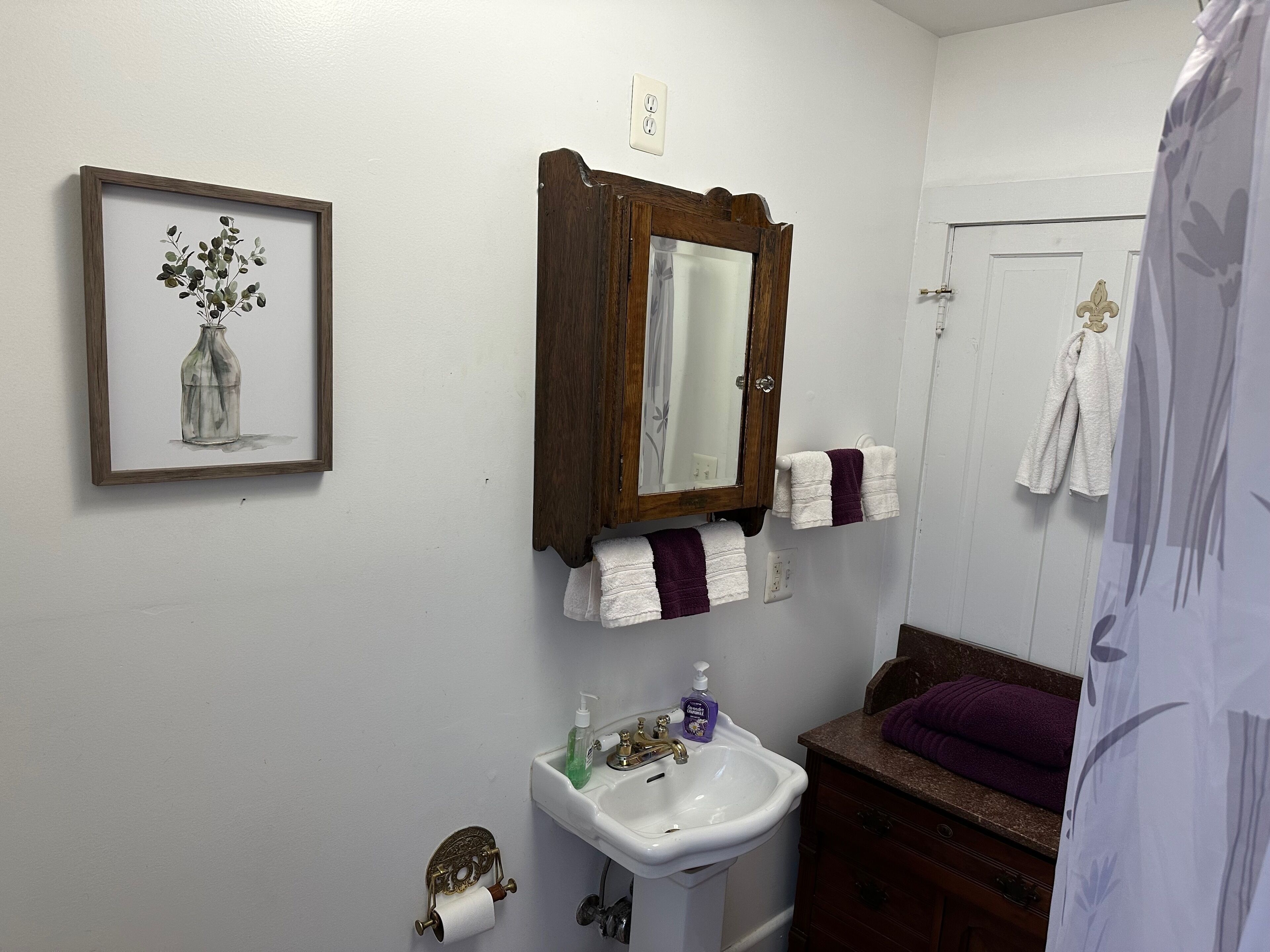 Casa de banho com uma pia branca, um espelho com moldura de madeira, uma obra de arte floral emoldurada e um porta-toalhas com toalhas brancas e roxas.