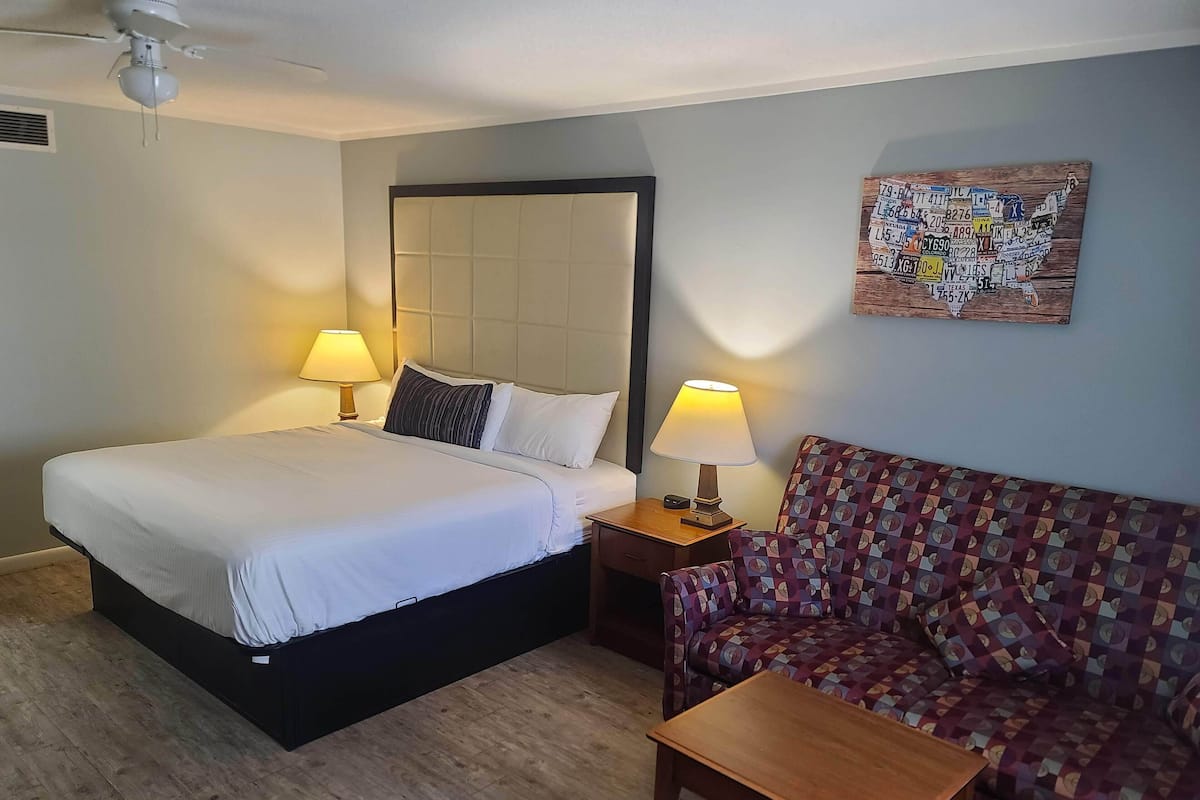 Quarto de hotel com cama, sofá, mesa pequena, duas luminárias de cabeceira e uma obra de arte na parede.