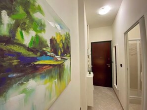 Um corredor com uma grande pintura abstrata à esquerda, uma porta no final e uma pequena mesa com um vaso.