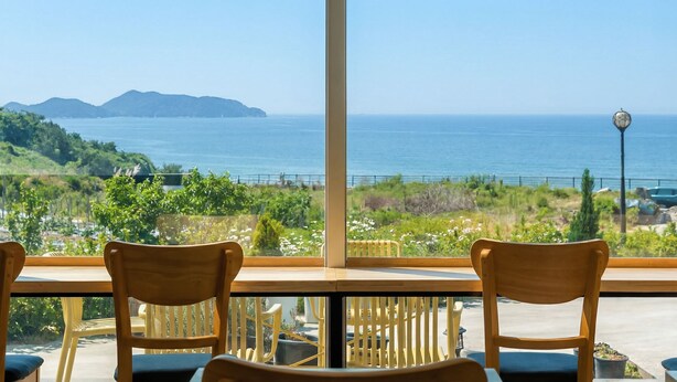 Vista de um quarto com grandes janelas para uma paisagem costeira, com uma mesa e cadeiras.
