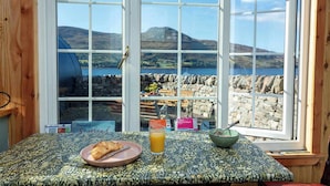 Quarto com uma grande janela com vista para uma paisagem costeira deslumbrante, uma mesa com um croissant, um copo de suco de laranja e uma tigela.