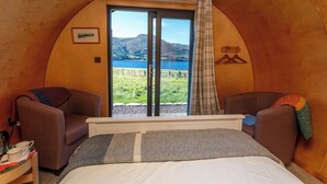 Quarto aconchegante com cama, duas poltronas e uma grande janela com vista panorâmica para um lago e montanhas.